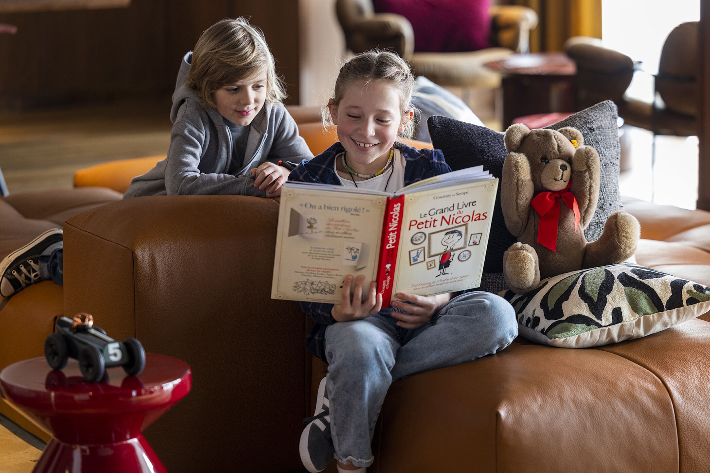 Duas crianças sentadas num sofá de couro marrom, lendo um livro intitulado 'Le Grand Livre de Petit Nicolas'.