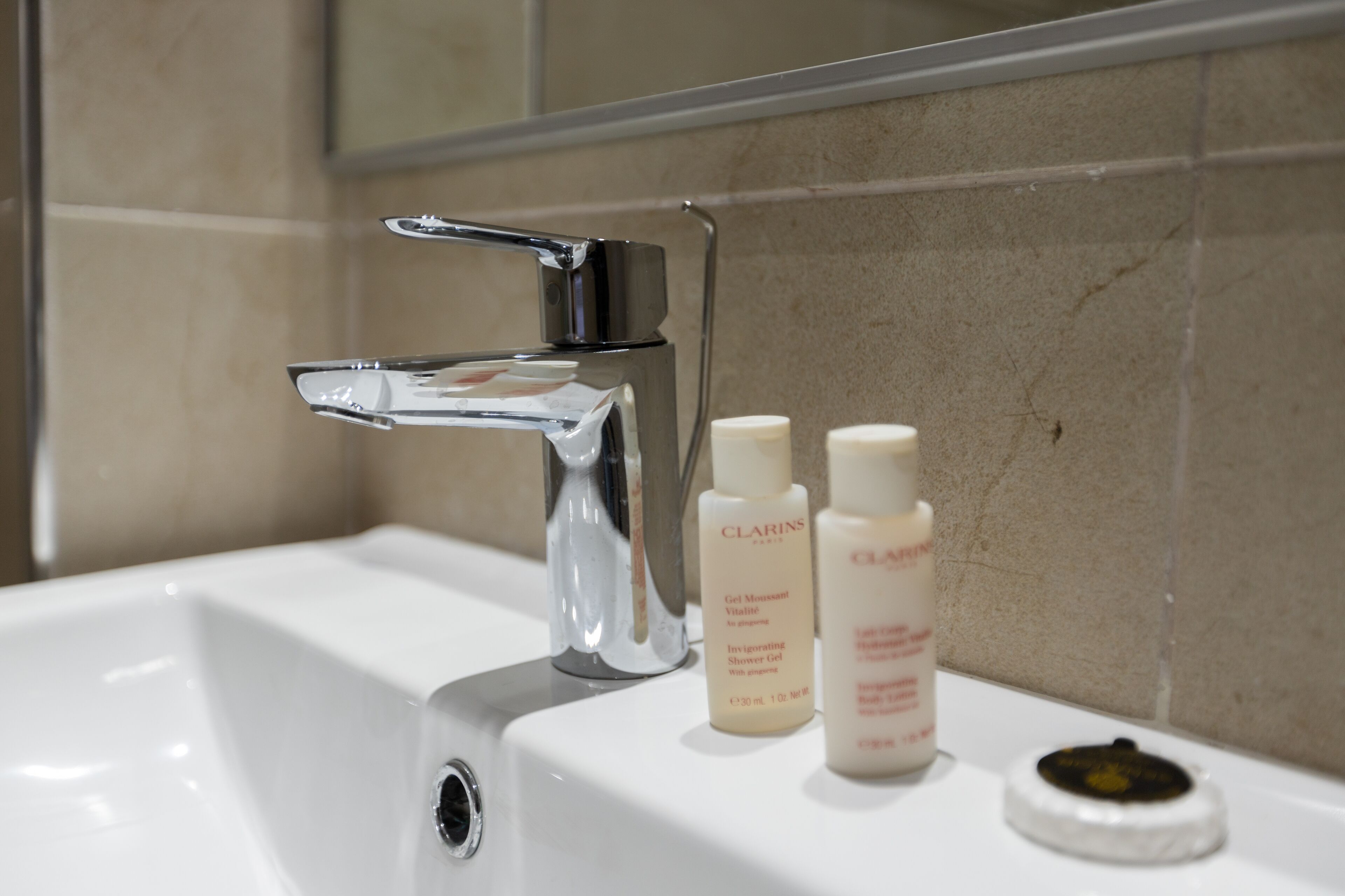 Uma pia de banheiro com torneira cromada, duas embalagens de gel de banho Clarins e um saboneteira.