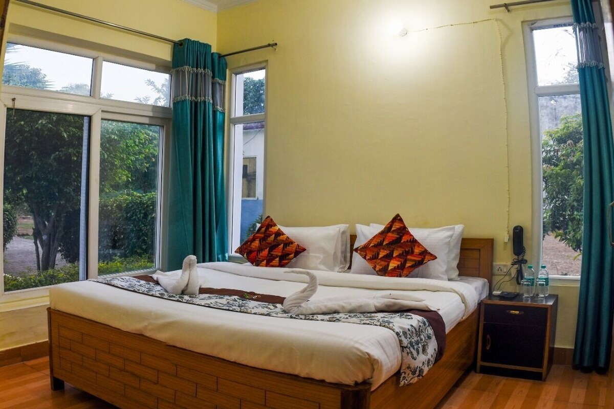 Quarto de hotel com cama de madeira, duas almofadas, mesa de cabeceira e janela com cortinas.