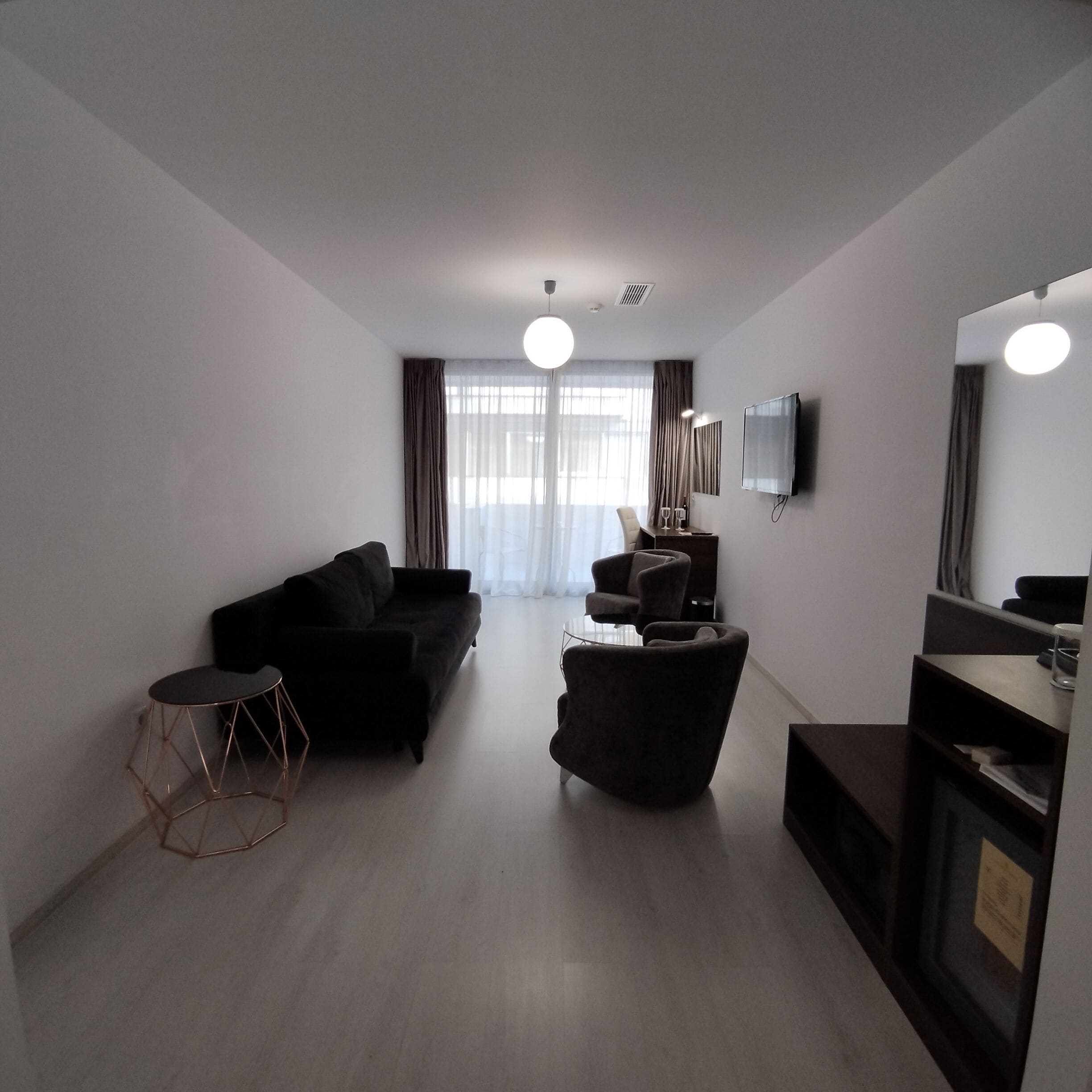 Quarto de hotel moderno com sofá preto, mesa de centro redonda, TV na parede e uma janela grande com cortinas.