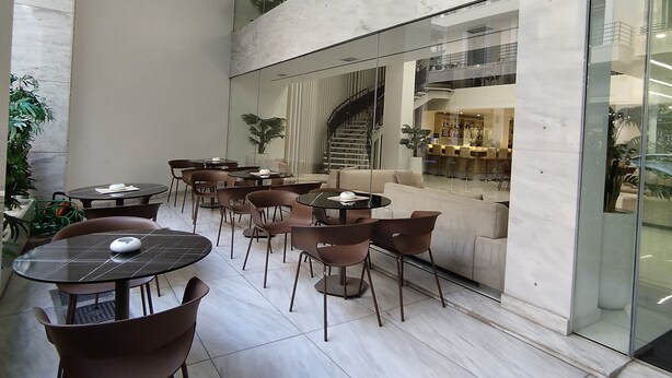 Um espaço amplo e arejado, com várias mesas e cadeiras, uma escadaria e vista para um restaurante ou café ao fundo.
