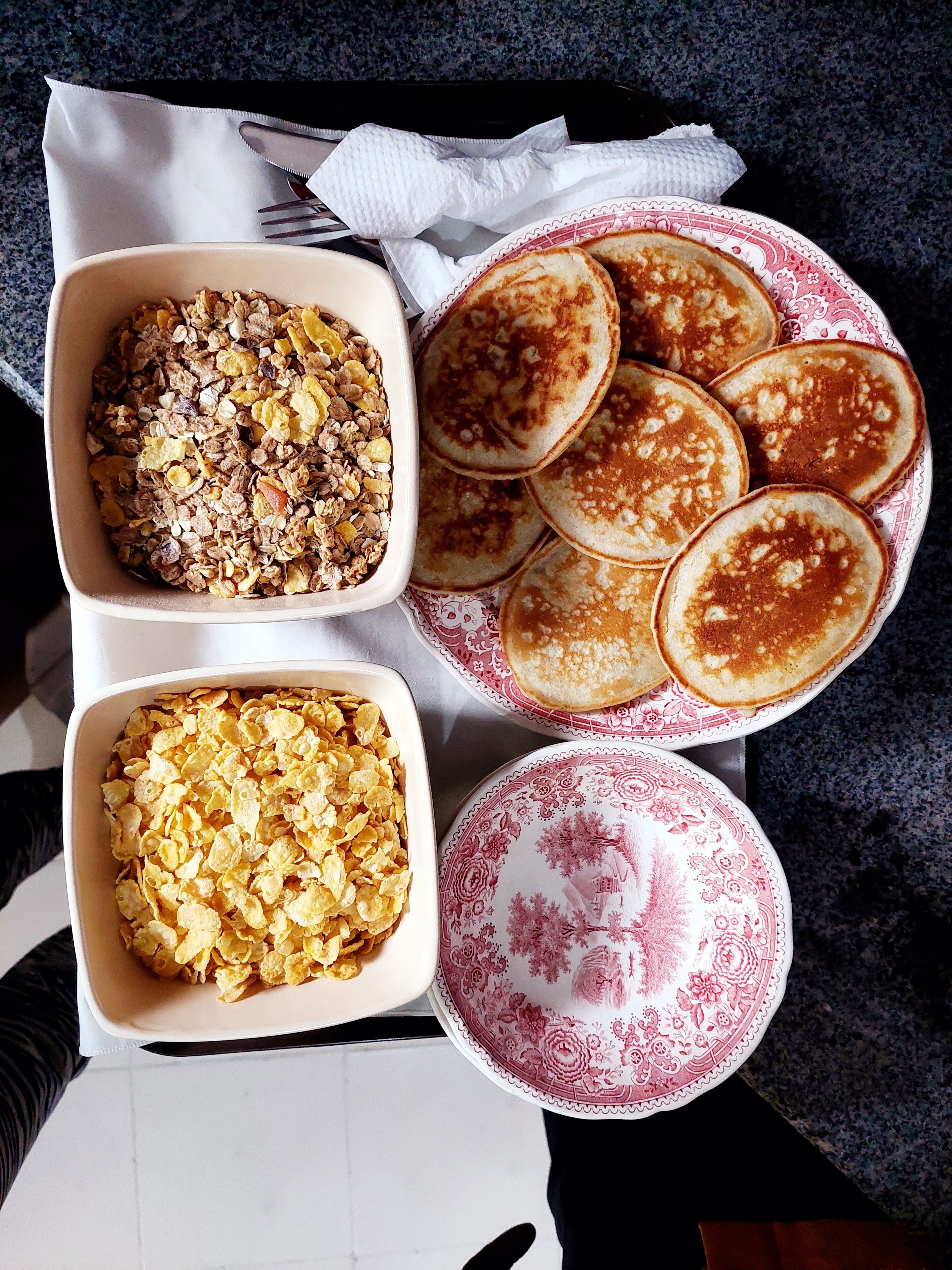 Bandeja de café da manhã com cereal, granola e panquecas sobre uma mesa.