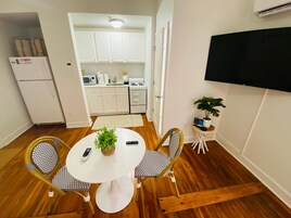 Cozinha compacta com armários brancos, micro-ondas, fogão e geladeira. Há uma área de refeições com mesa branca e duas cadeiras, um vaso de planta e uma TV fixada na parede.