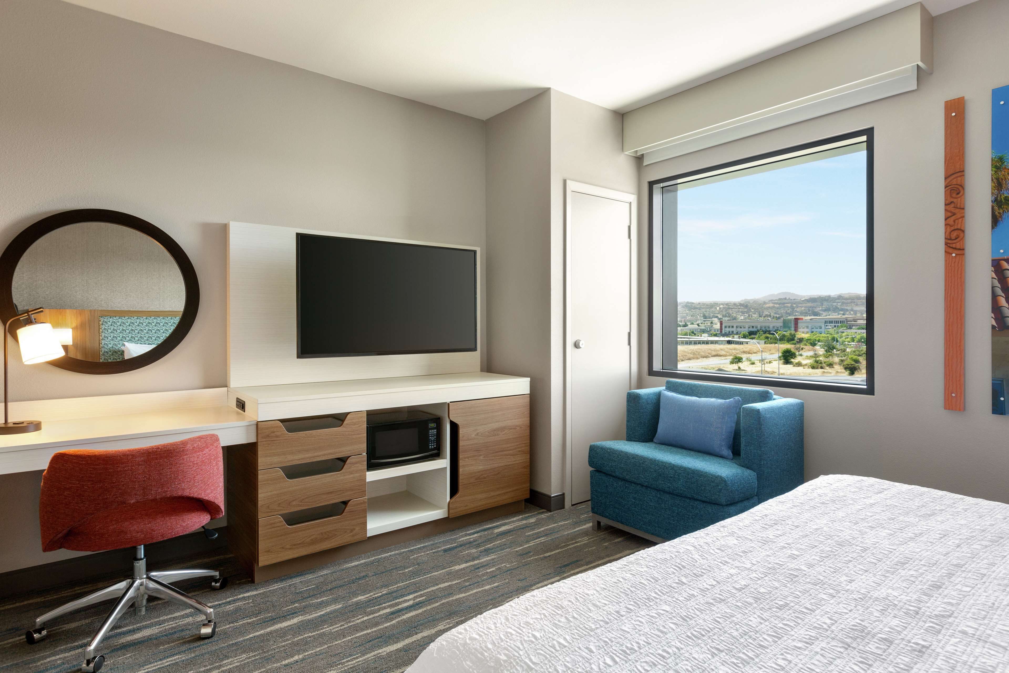 Quarto de hotel com cama, escrivaninha com cadeira, televisão na parede, janela grande com vista e painel de madeira na parede.