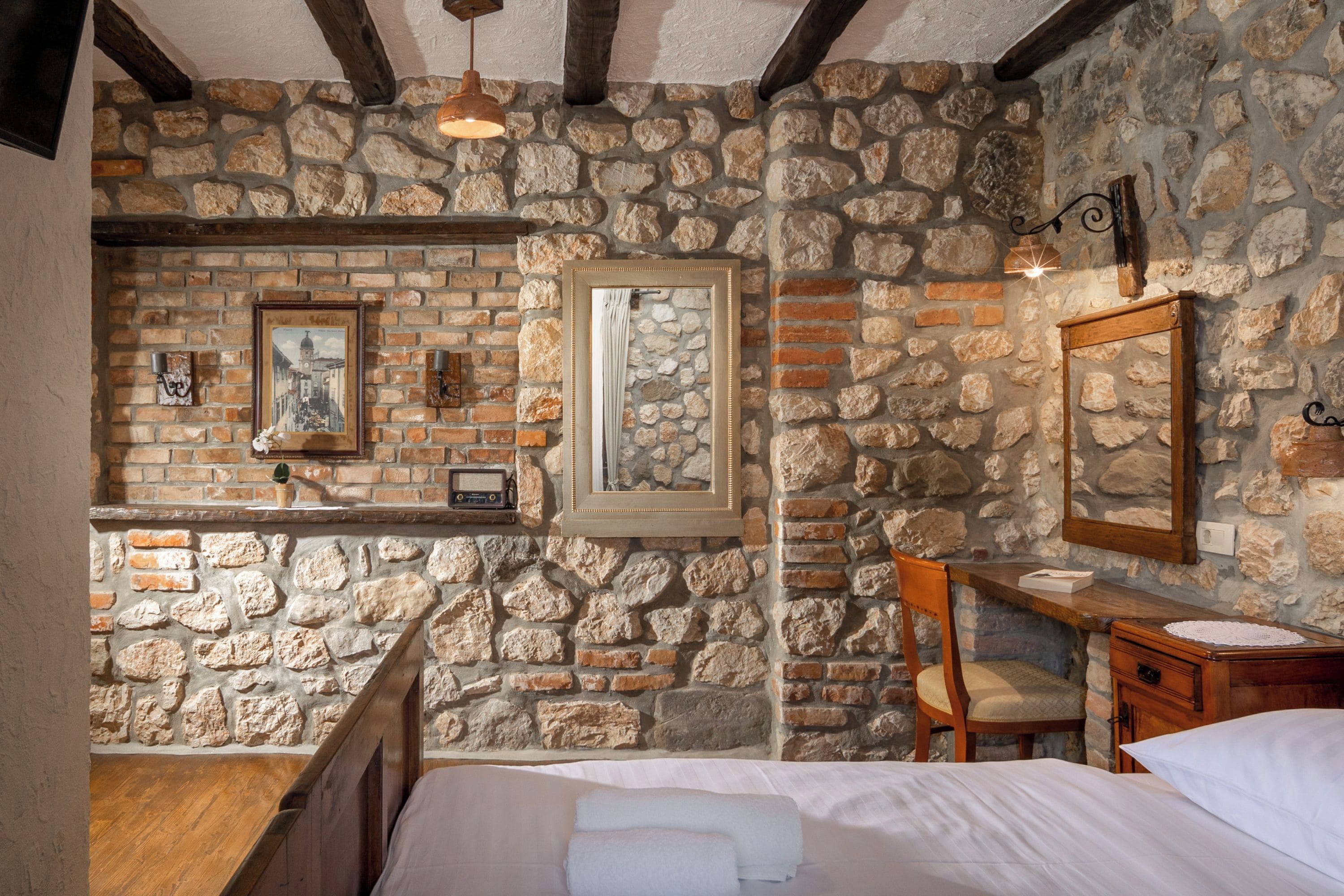 Quarto rústico com paredes de pedra, uma escrivaninha de madeira, uma cadeira, uma cama e uma janela.