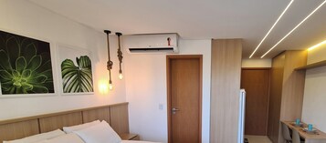 Quarto de hotel com cama, ar condicionado fixado na parede, porta e pequena mesa com xícara e pires.