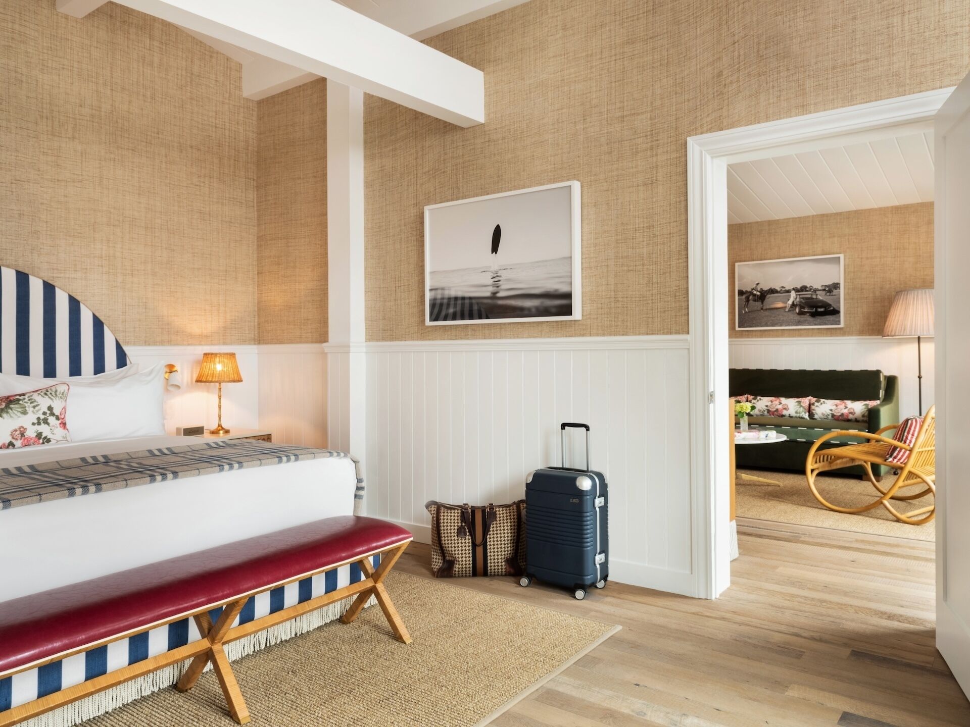 Quarto de hotel com cama, um banco, malas e vista para outro quarto.