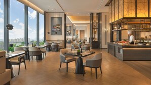 Um restaurante moderno com um chef preparando alimentos, grandes janelas com vista para a cidade e um amplo espaço de refeições com pé direito alto e luz natural.