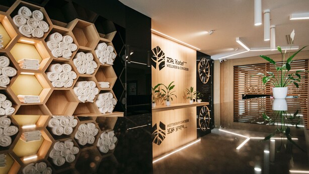 Uma área de recepção de hotel com design moderno, apresentando uma exibição hexagonal de toalhas em madeira, um balcão de recepção e um vaso de planta.