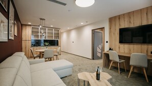 Uma sala de estar moderna com sofá, área de jantar com mesa e cadeiras, cozinha ao fundo e TV fixada em uma parede de madeira.