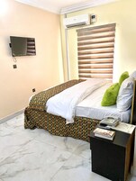Quarto com cama, televisão fixada na parede, ar condicionado e janela com persianas.