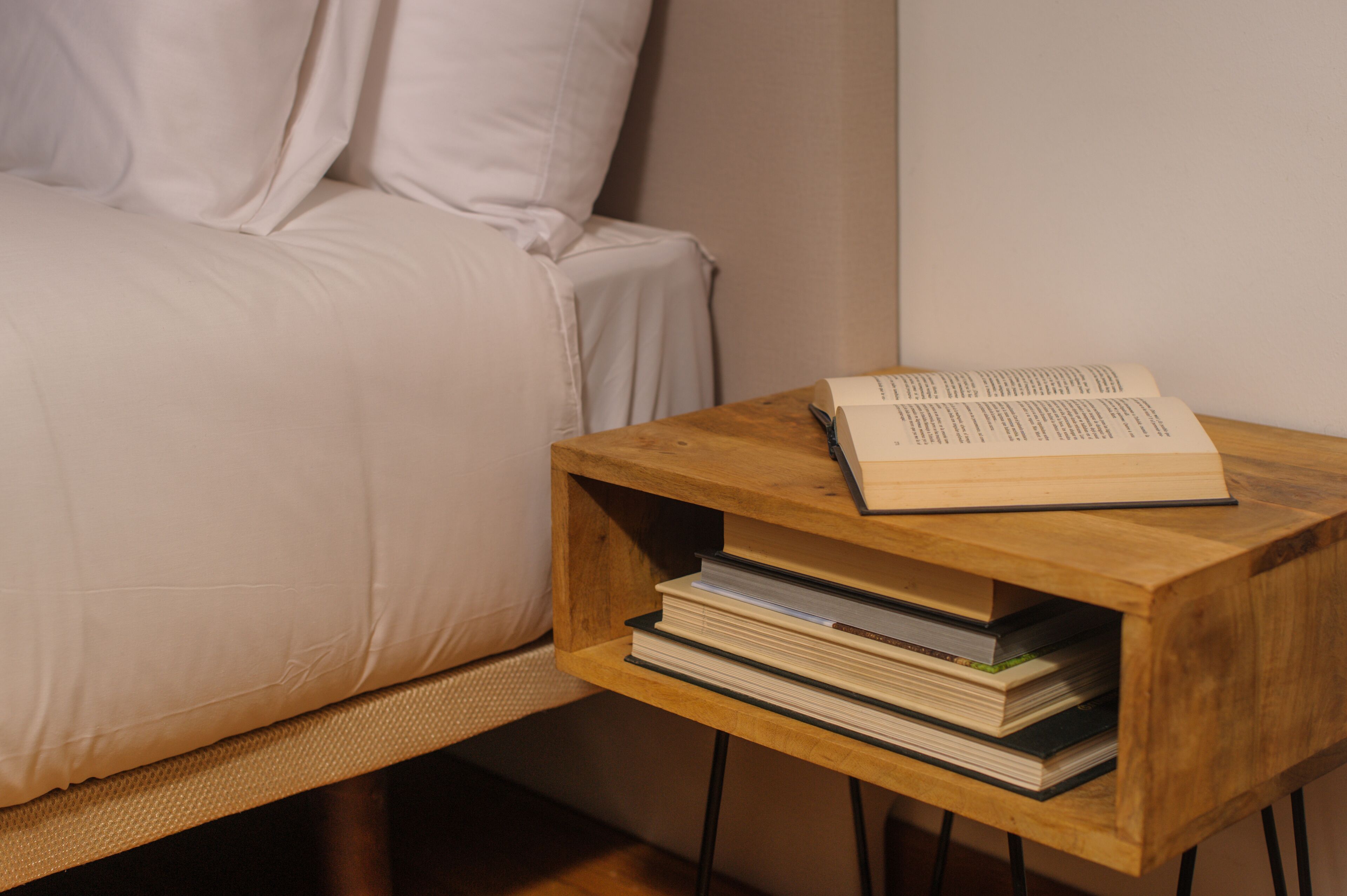 Uma cama bem arrumada com lençóis brancos, uma mesa de cabeceira de madeira com um livro e uma pilha de livros, e um cesto trançado debaixo da cama.