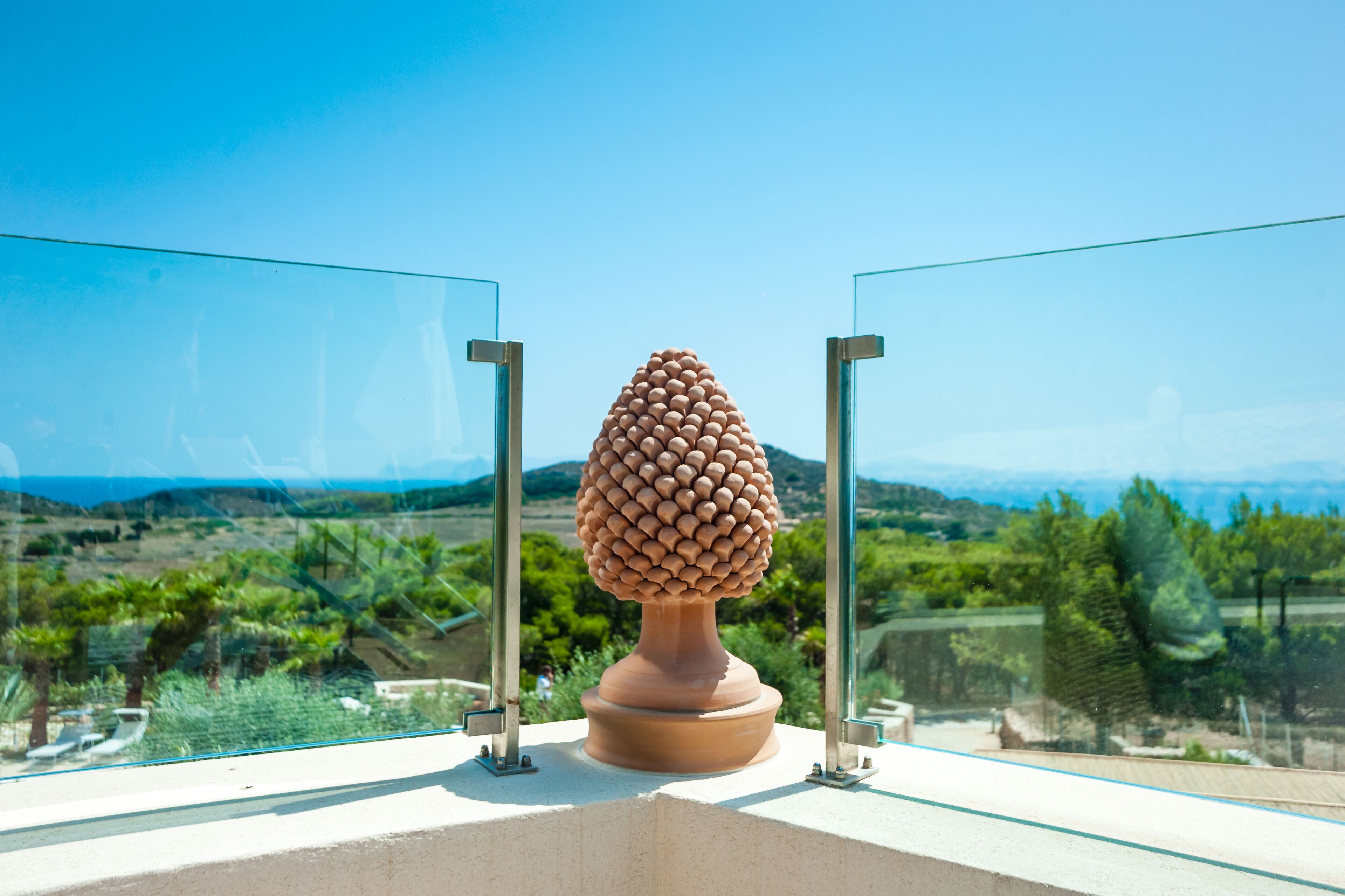 Escultura em terracota em uma varanda com grades de vidro, com vista para uma paisagem verde e colinas ao longe.