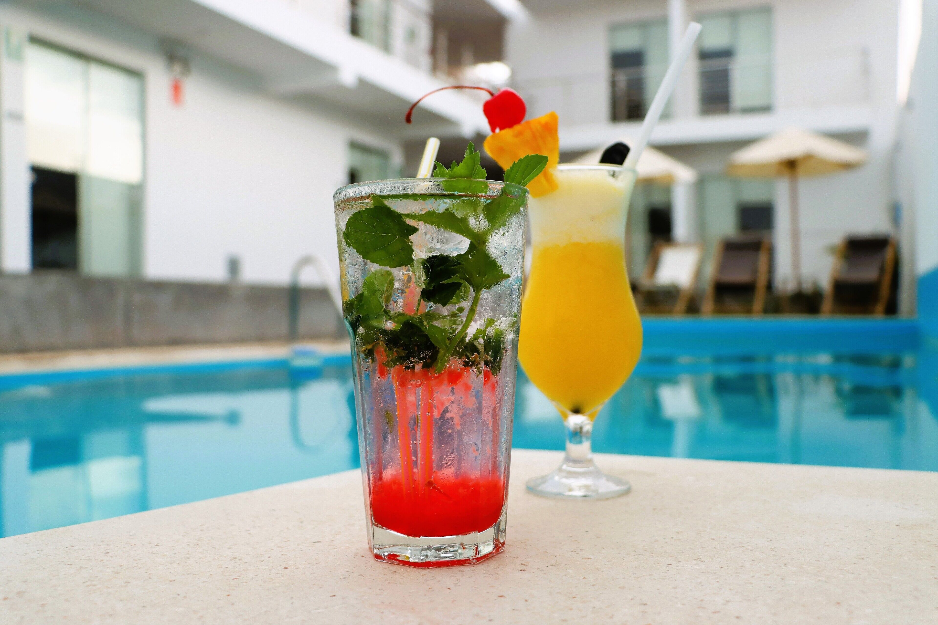 Dois cocktails numa mesa junto a uma piscina, com um edifício ao fundo.