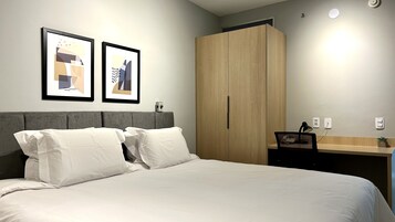 Cama bem arrumada com lençóis brancos, cabeceira estofada cinza e duas obras de arte abstratas emolduradas na parede.