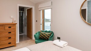 Um quarto com cômoda de madeira, uma poltrona verde, uma cama com manta branca, uma janela com vista e um espelho redondo na parede.