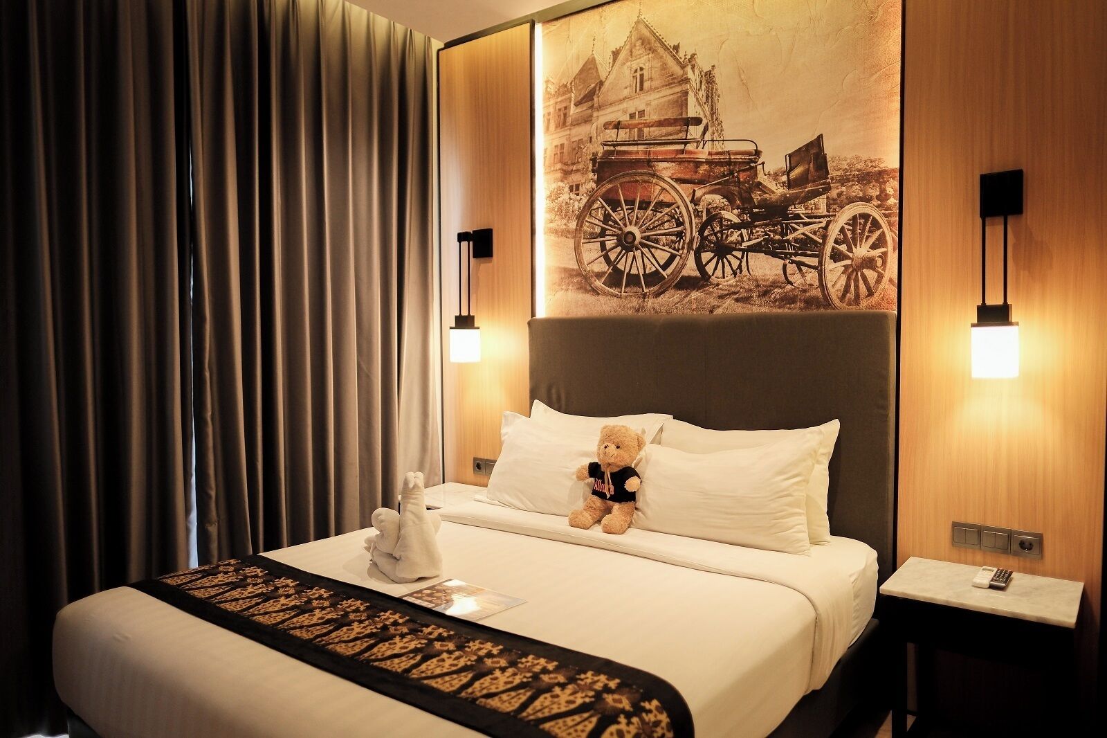 Quarto de hotel com uma cama grande, um ursinho nos travesseiros, uma televisão na parede e um quadro de uma carruagem puxada por cavalos.