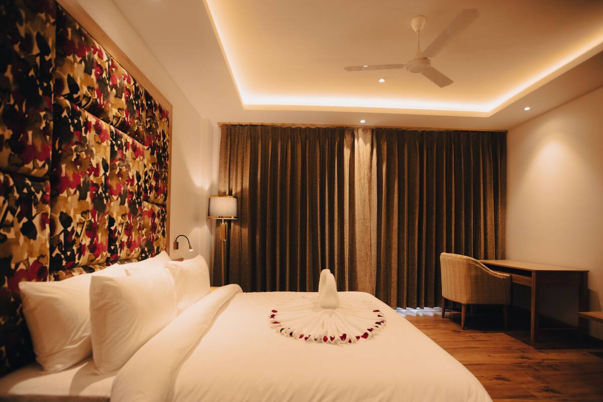 Quarto de hotel com cama, escrivaninha, cadeira, ventilador de teto e parede com padrão floral.