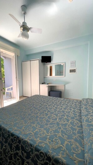 Um quarto com cama, escrivaninha, cadeira, guarda-roupa, ventoinha no teto e televisão.