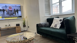Sala de estar com um sofá verde, uma mesa de centro de vidro e uma TV exibindo notícias.