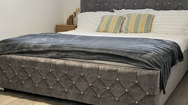 Uma cama com cabeceira estofada, uma mesinha de cabeceira com abajur e uma janela com cortinas.