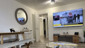 Uma sala de estar moderna com um banco de madeira, uma mesa de centro de vidro e uma televisão de tela plana exibindo notícias.