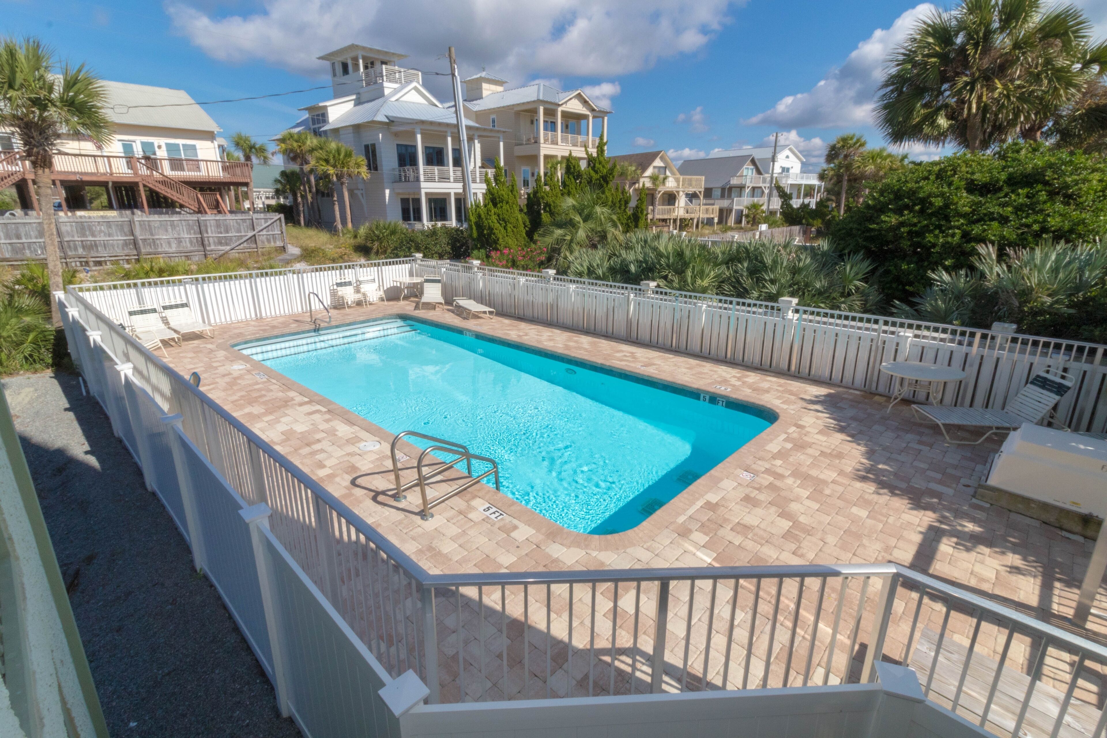 Piscina retangular com deck branco e escada de metal, cercada por uma cerca branca.