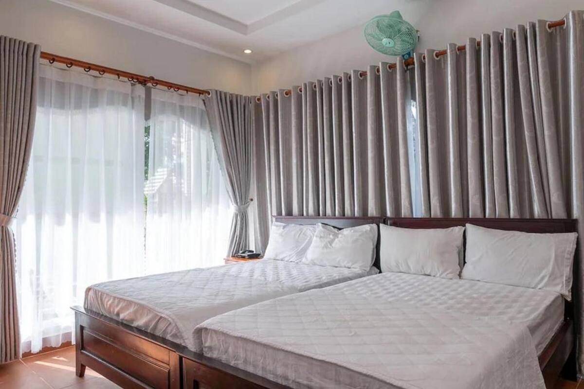 Quarto de hotel com duas camas, cabeceira de madeira, ventoinha no teto e cortinas leves.