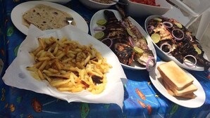 Mesa posta com diversos pratos, incluindo peixe grelhado, batatas fritas e pão, com toalha de mesa azul estampada com motivos de peixe.
