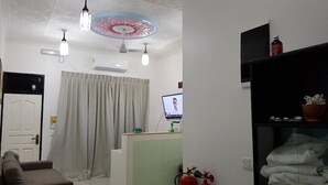 Quarto com ventoinha no teto, televisão fixada na parede, extintor de incêndio, porta e janela com cortinas.