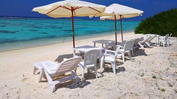 Um cenário à beira-mar com cadeiras e mesas de lounge brancas sob guarda-sóis, com vista para um mar azul cristalino.