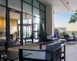 Área de estar externa com móveis modernos, entrada de vidro e vista para a paisagem urbana.