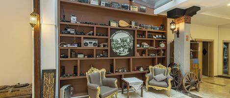 Um hall com teto alto, telhado de vidro e parede decorada com rádios antigos e uma grande fotografia de família. Há duas poltronas elaboradas e uma pequena mesa.