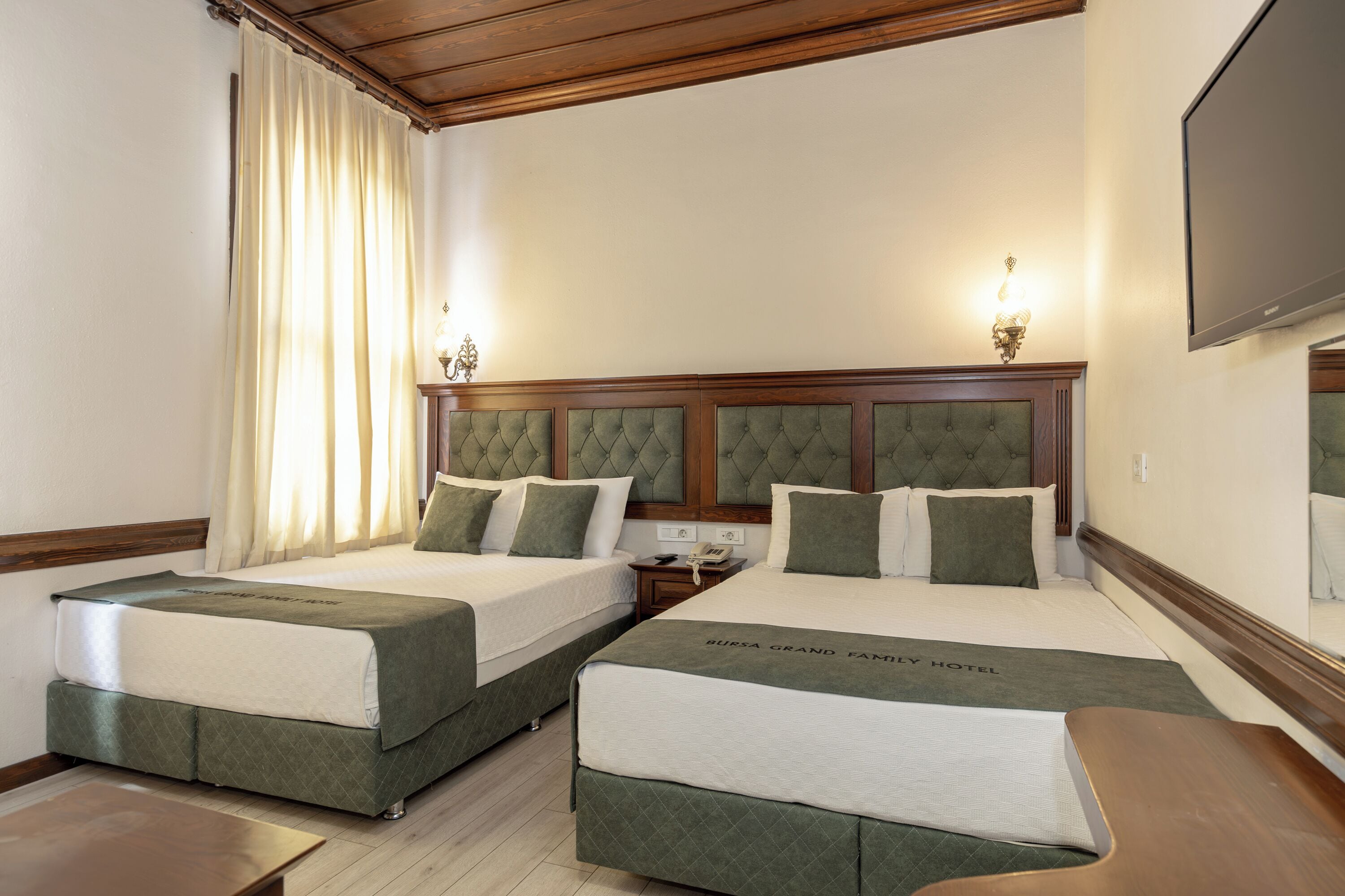 Quarto de hotel com duas camas, cabeceira de madeira, mesa de cabeceira, luminária e televisão.