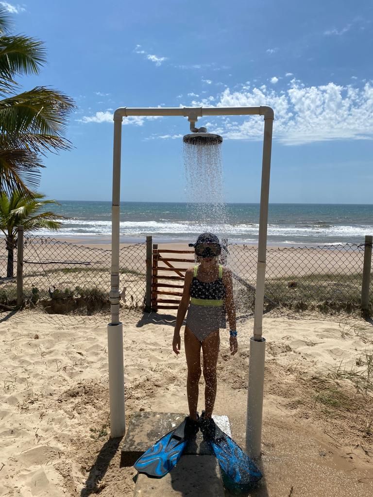 Uma pessoa em pé sob uma estrutura de chuveiro na praia, com um cercado e palmeiras ao fundo.