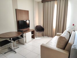 Quarto de hotel moderno com televisão de tela plana, mesa de jantar com cadeiras, sofá e janela com cortinas.