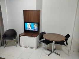 Quarto com televisão fixada em um painel de madeira, um armário branco, uma mesa de madeira redonda e quatro cadeiras pretas.