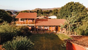 Um edifício grande, de dois andares, com telhado de telhas vermelhas, rodeado por vegetação exuberante e colinas ao fundo.