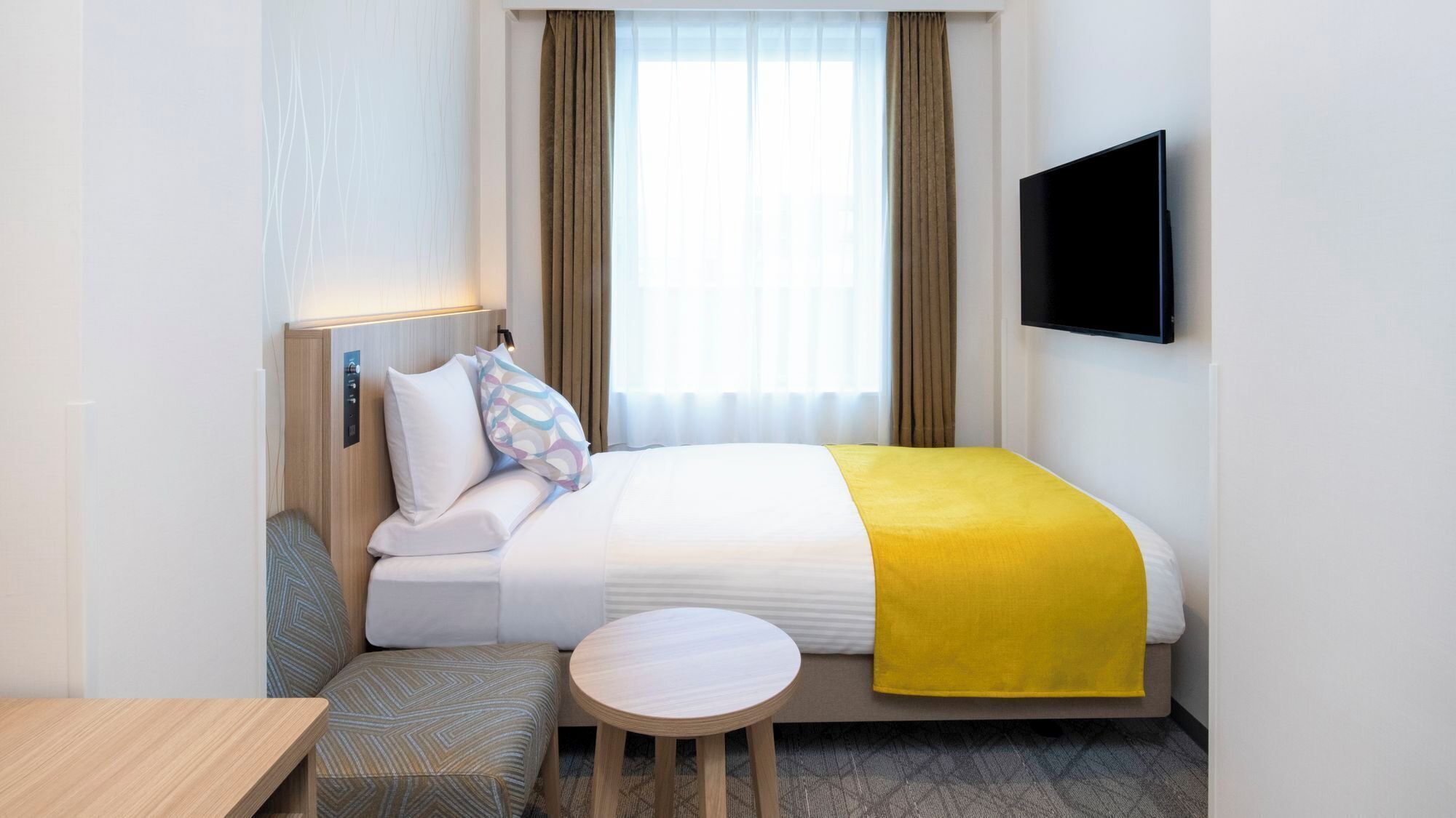 Quarto de hotel com cama, mesa de cabeceira, mesinha redonda, televisão na parede e janela com cortinas.