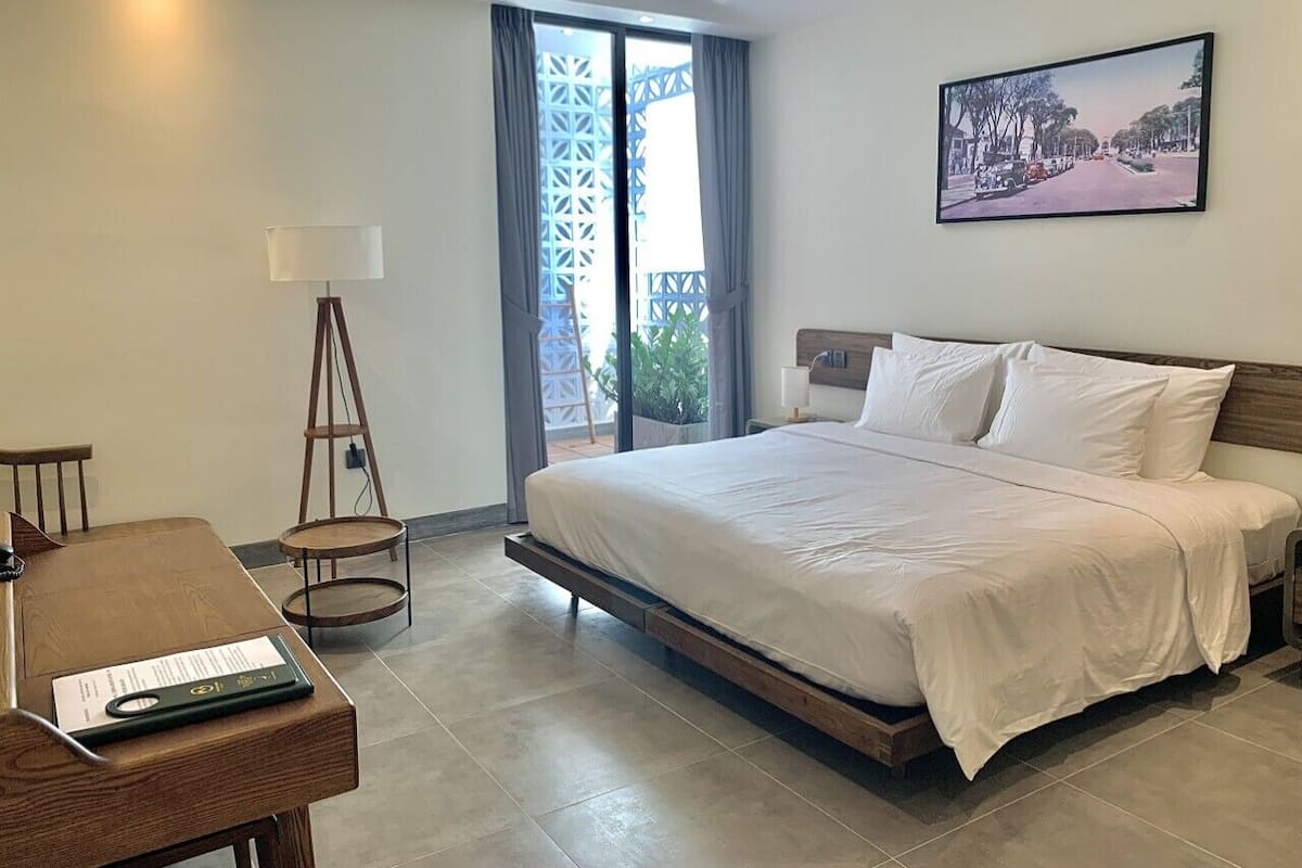 Quarto de hotel com cama, escrivaninha, cadeira, abajur e vista para o exterior pela janela.