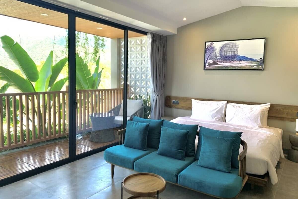 Quarto com cama, sofá, mesa pequena e varanda com vista para o verde.