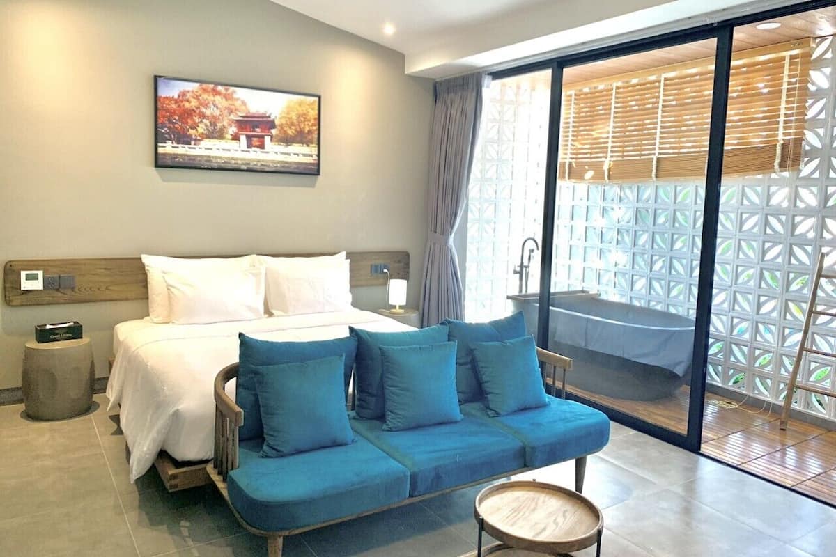 Quarto de hotel moderno com cama, sofá azul, pequena mesa redonda e grande janela com vista para piscina e área externa.