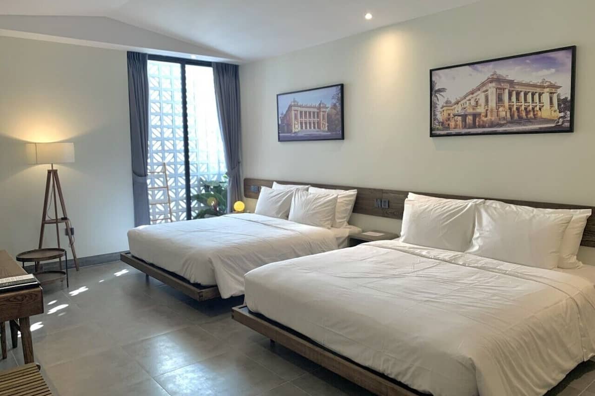 Quarto de hotel com duas camas, uma escrivaninha com um laptop, uma lâmpada e uma janela com vista para um prédio.