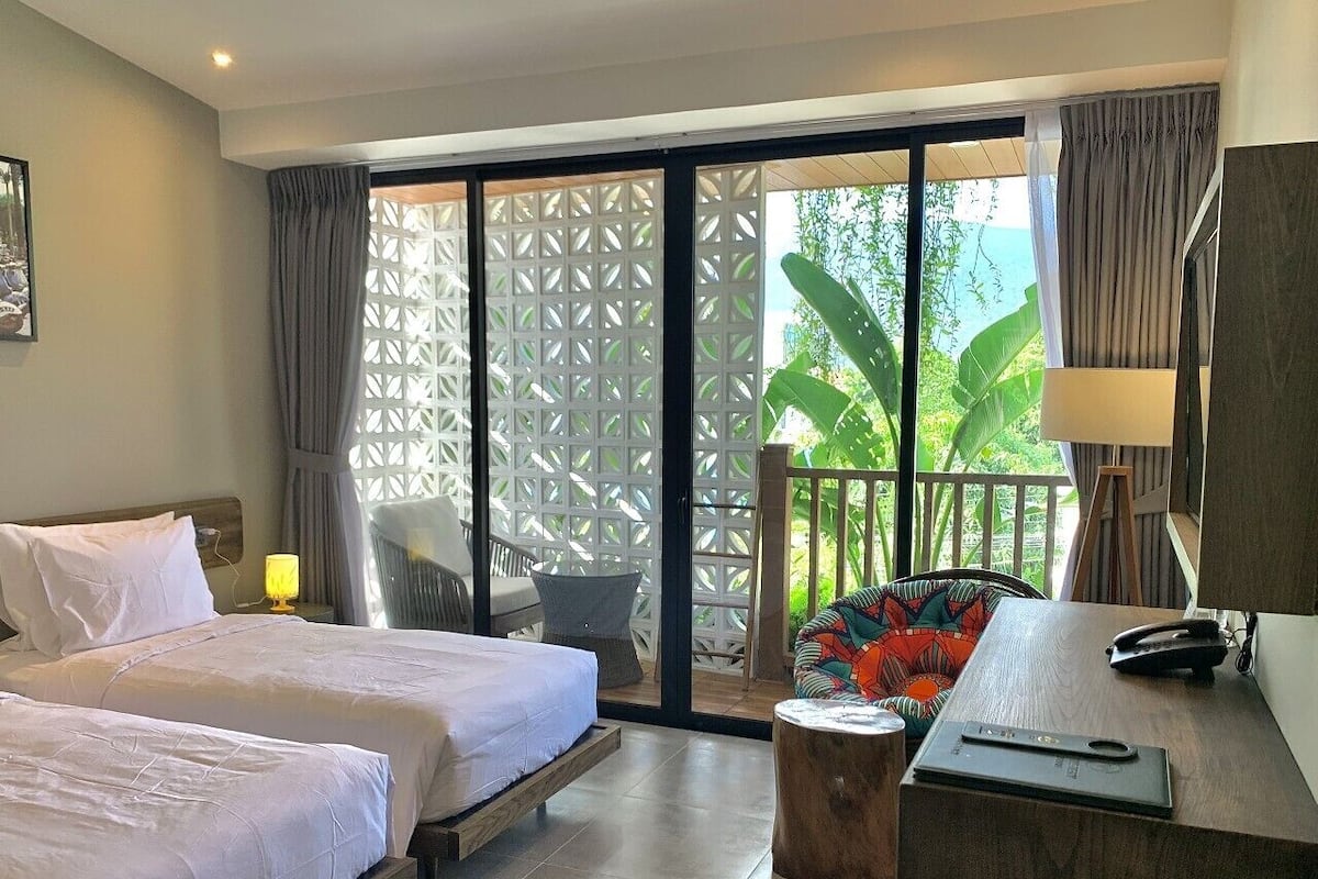 Quarto de hotel com duas camas, uma mesa com telefone, uma cadeira e varanda com vista para vegetação.