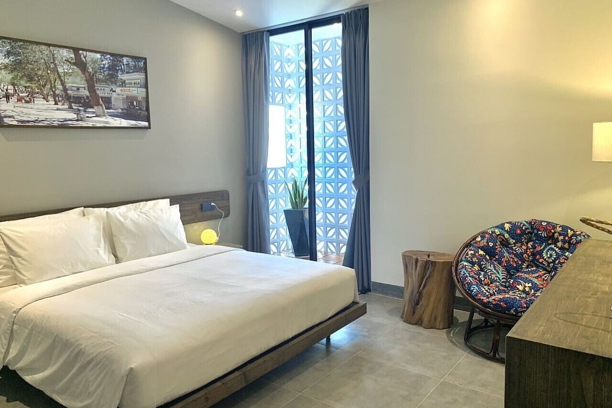 Quarto de hotel moderno com uma cama grande, uma poltrona, uma mesinha e vista através de uma janela com padrão geométrico.