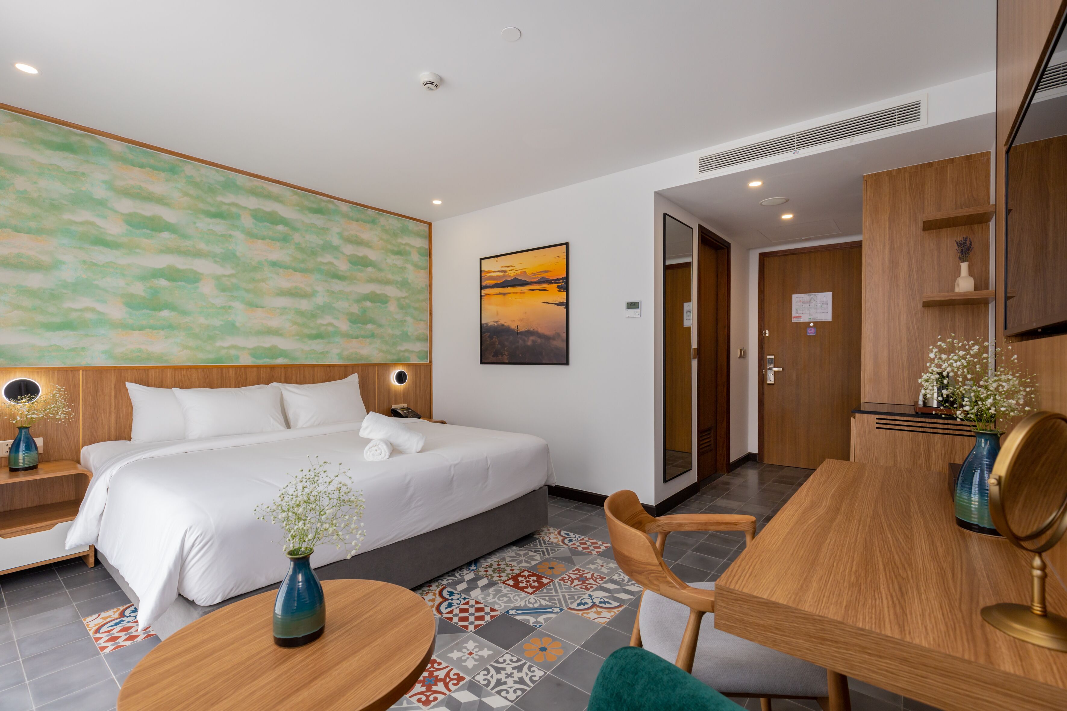 Quarto de hotel com uma cama grande, uma escrivaninha de madeira, uma cadeira, um vaso com flores e um quadro na parede.
