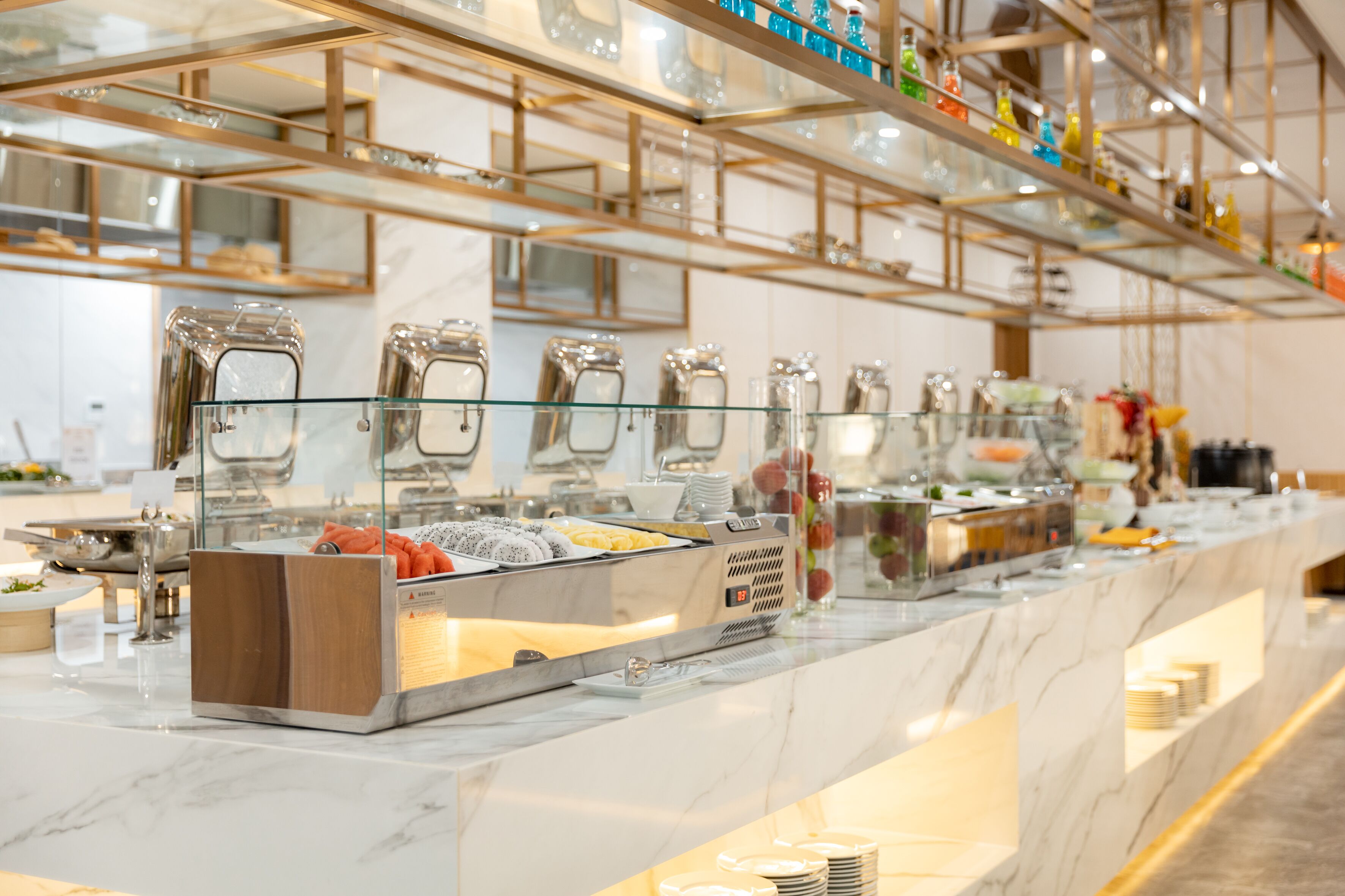 Buffet com vários recipientes de vidro para alimentos, bancada de mármore e suporte de metal com garrafas.