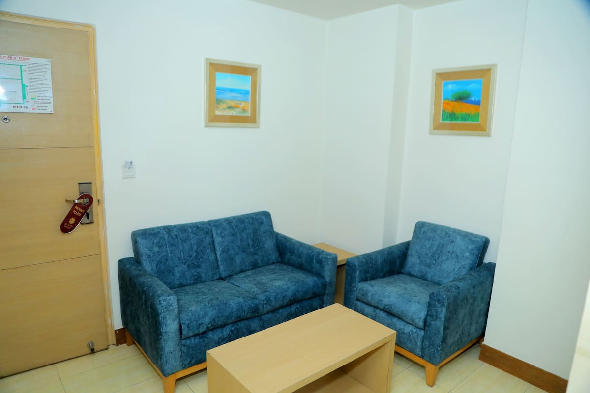 Quarto com dois sofás azuis, uma mesa de madeira, uma porta de madeira com um alarme de incêndio e duas molduras com quadros na parede.