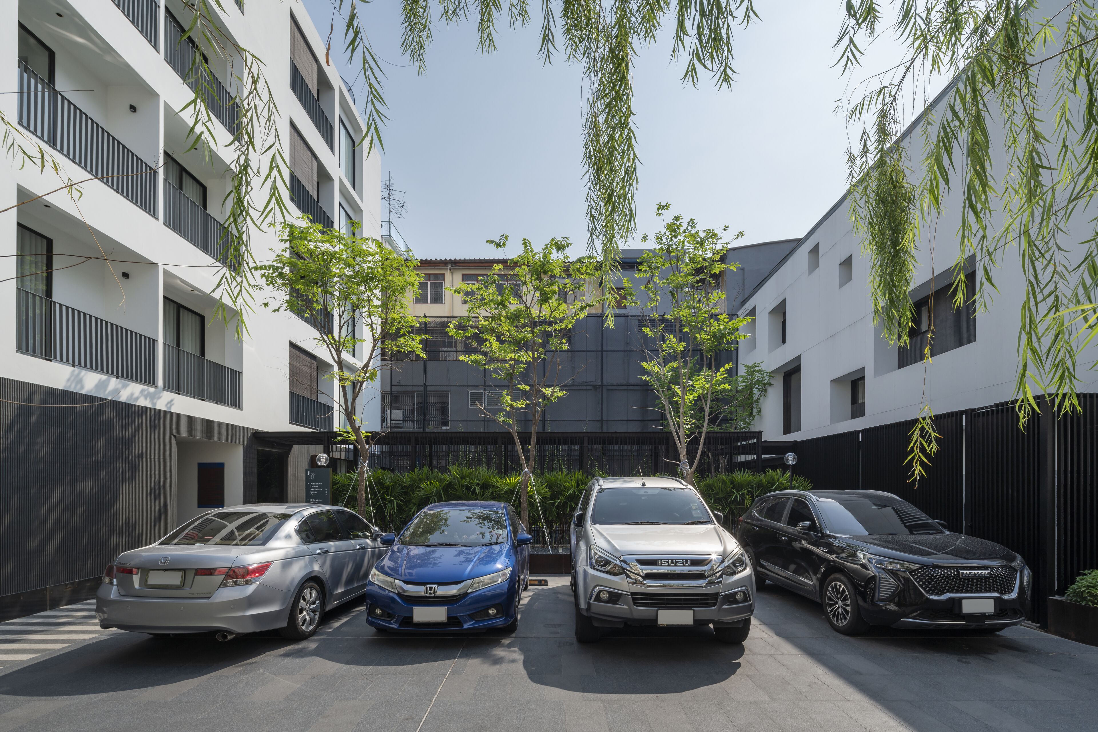 Uma área de estacionamento com quatro carros estacionados em frente a um edifício moderno com varandas e uma árvore ao centro.
