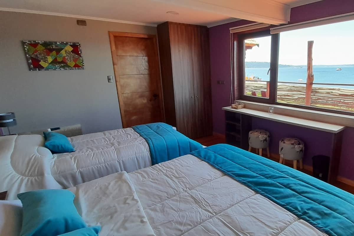Quarto com duas camas, janela com vista para o mar, guarda-roupa de madeira e uma decoração de parede colorida.